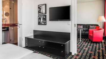 Quarto de hotel moderno com cama, televisão preta, escrivaninha com gavetas, cadeira vermelha e carpete estampado.