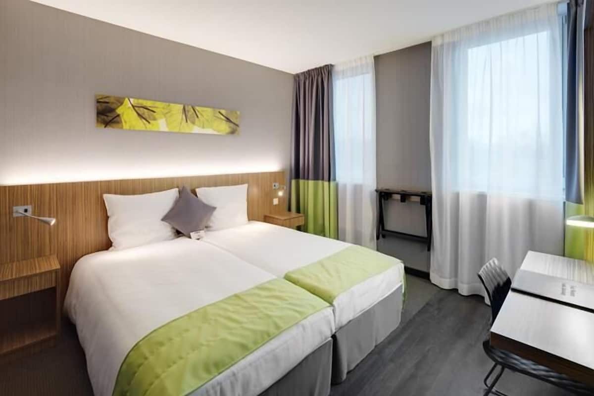 Quarto de hotel com uma cama grande, uma escrivaninha e uma cadeira. Há um quadro na parede e uma janela com cortinas.