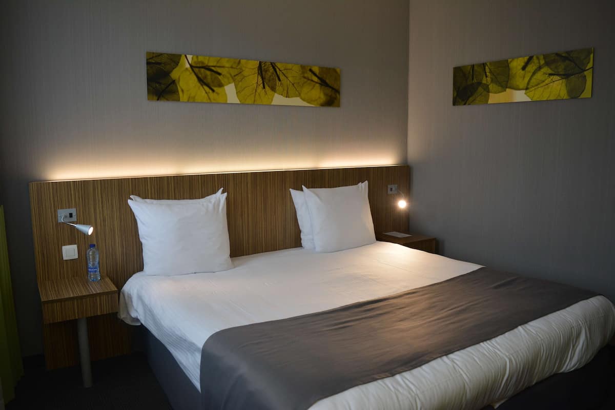 Quarto de hotel com uma cama grande, duas almofadas brancas, cabeceira de madeira, mesa de cabeceira com abajur e duas obras de arte penduradas na parede.