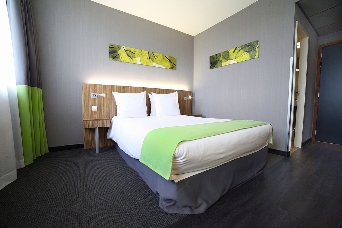 Quarto de hotel com cama, mesa de cabeceira, cortina verde e duas obras de arte na parede.