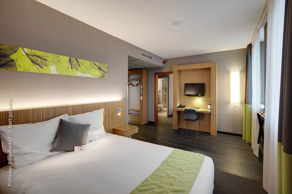 Quarto de hotel com cama, escrivaninha, televisão e banheiro visível através de uma porta aberta.
