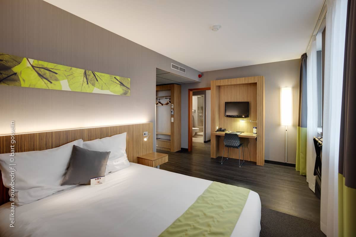 Quarto de hotel com uma cama grande, uma escrivaninha, uma televisão e uma casa de banho visível através de uma porta aberta.