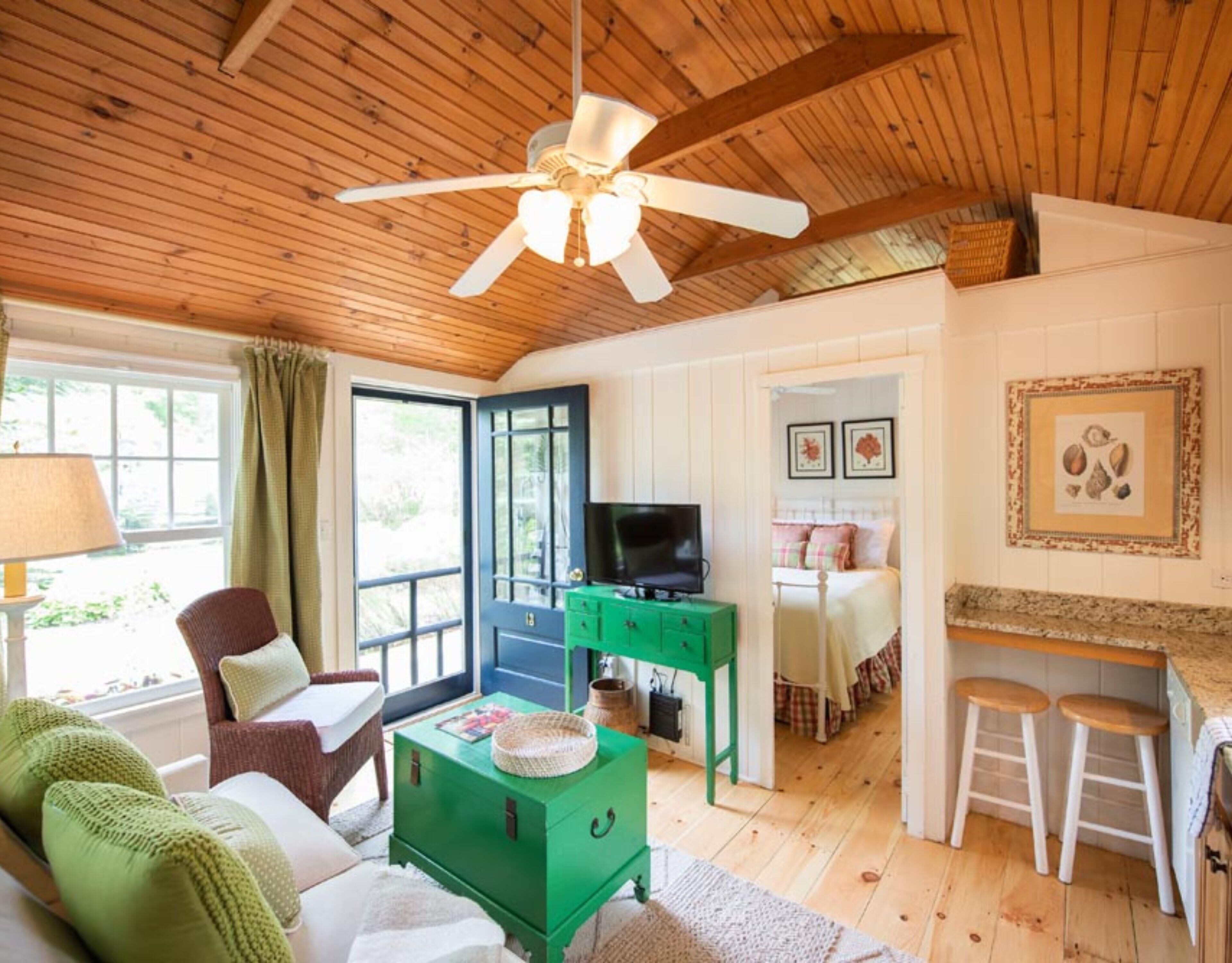 Quarto aconchegante com teto de madeira, uma escrivaninha verde, televisão, cama e janela com cortinas.