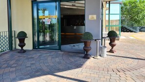 Entrada de hotel com porta de vidro, placa com a inscrição "benvenuti!" e plantas em vasos.