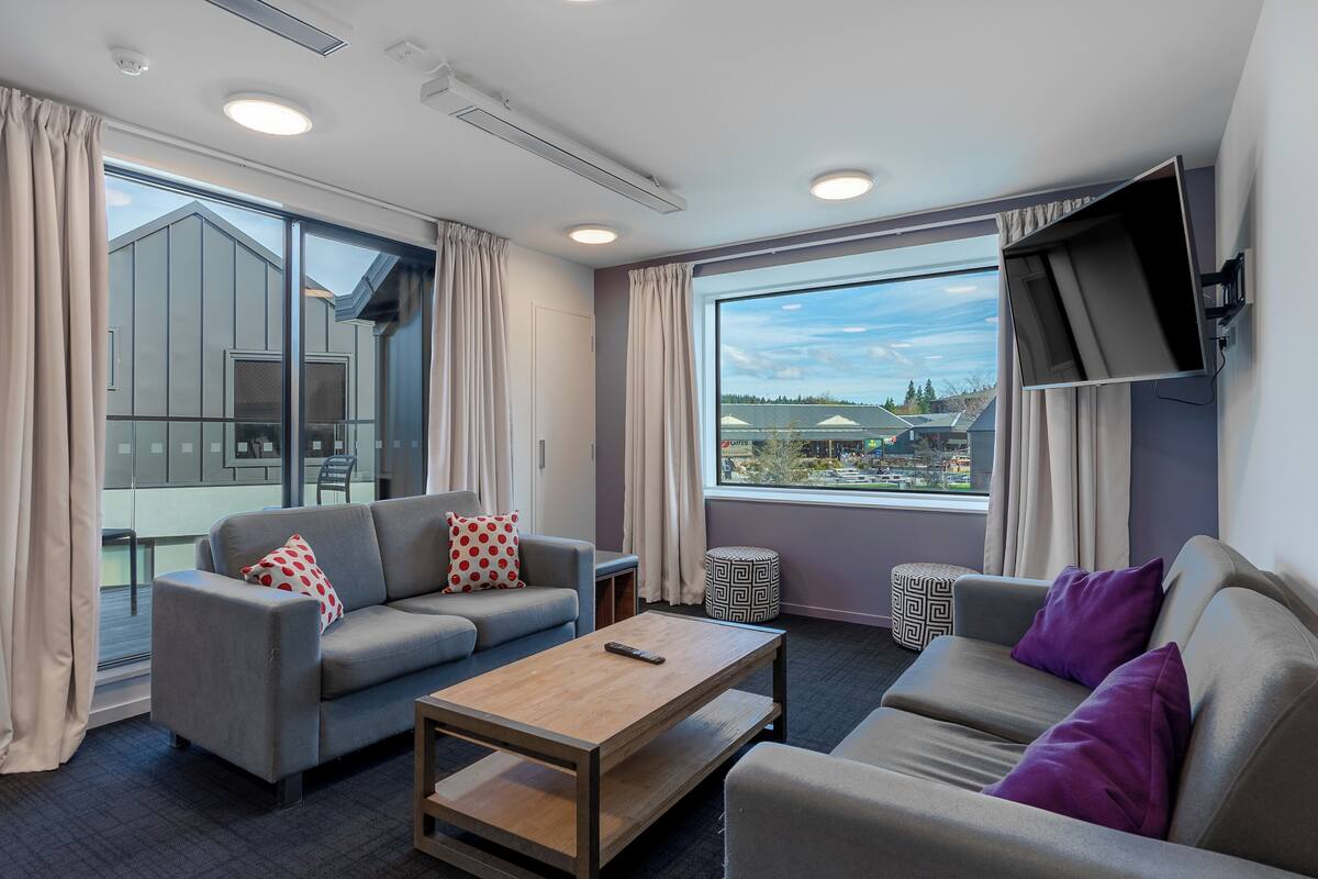 Quarto de hotel moderno com sofá cinza, mesa de centro de madeira, televisão fixada na parede e uma grande janela com vista para a cidade.
