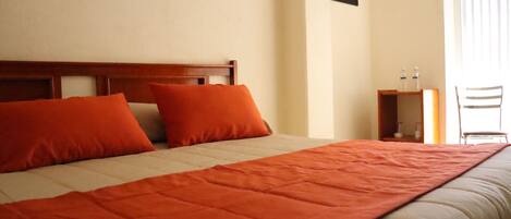 Cama bem arrumada com manta e travesseiros vermelhos, cabeceira de madeira e pequena mesa de cabeceira com abajur.