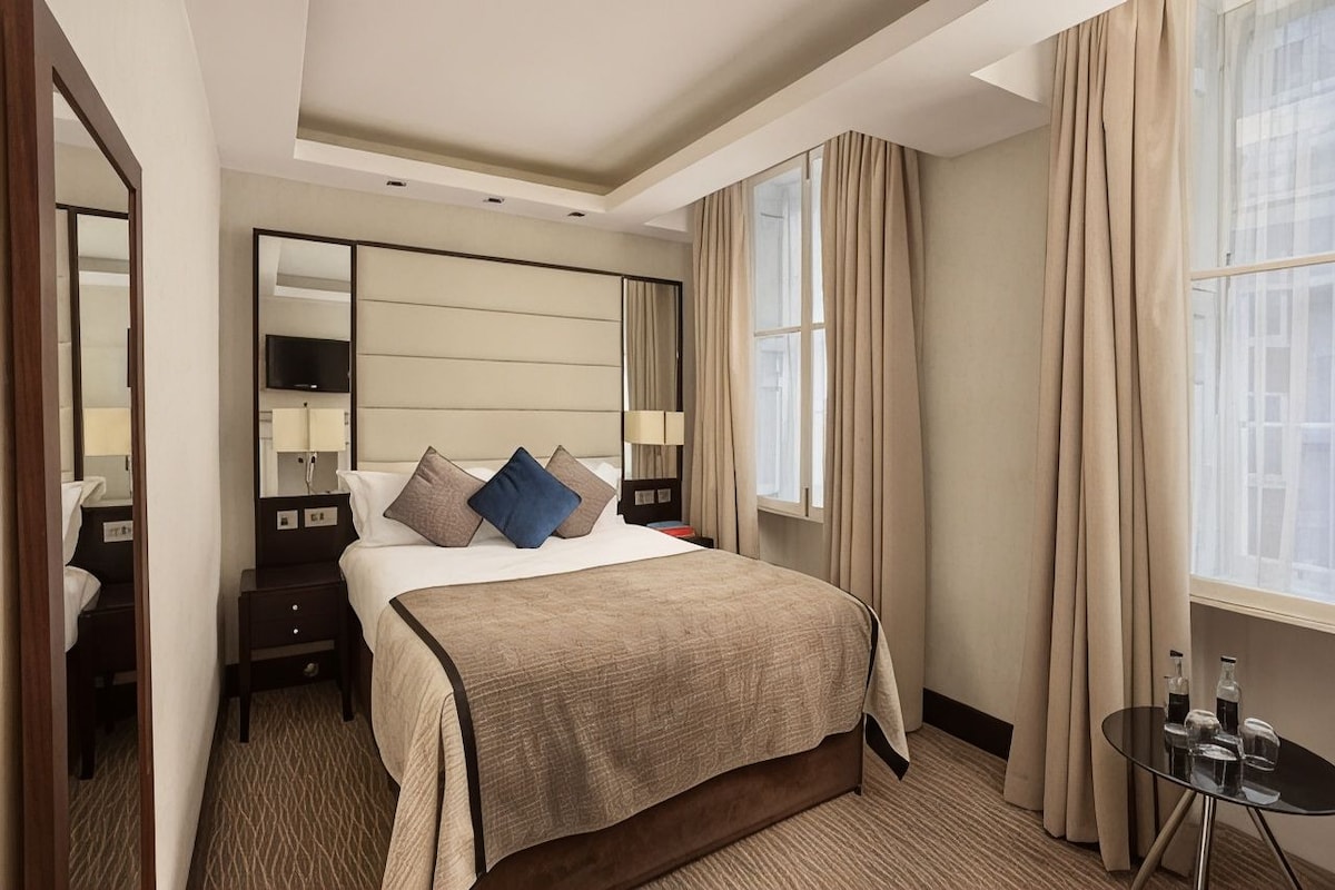 Quarto de hotel com cama grande, mesa de cabeceira, espelho, televisão e janela com cortinas.