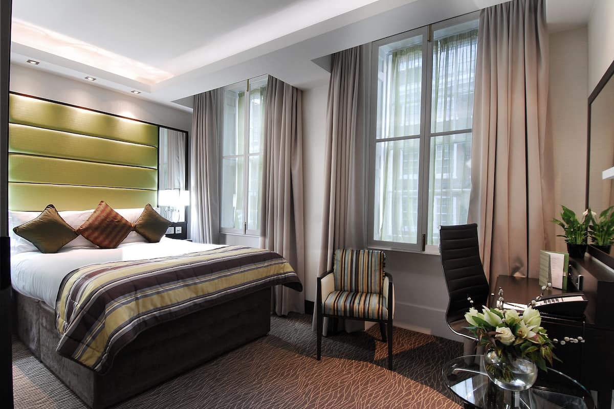 Quarto de hotel com cama, uma cadeira, escrivaninha e janela.