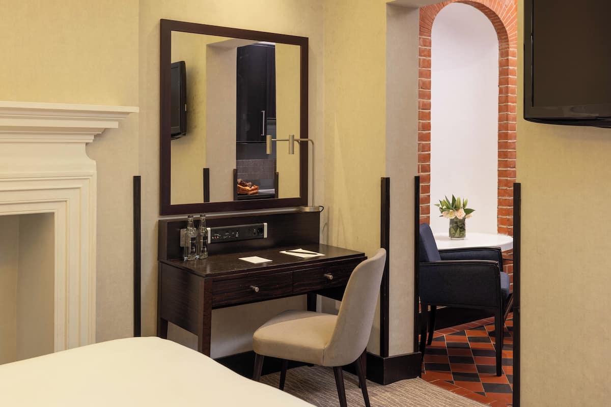 Quarto de hotel com uma cama, uma escrivaninha com cadeira, um espelho, uma televisão na parede e uma mesinha com um vaso de flores.