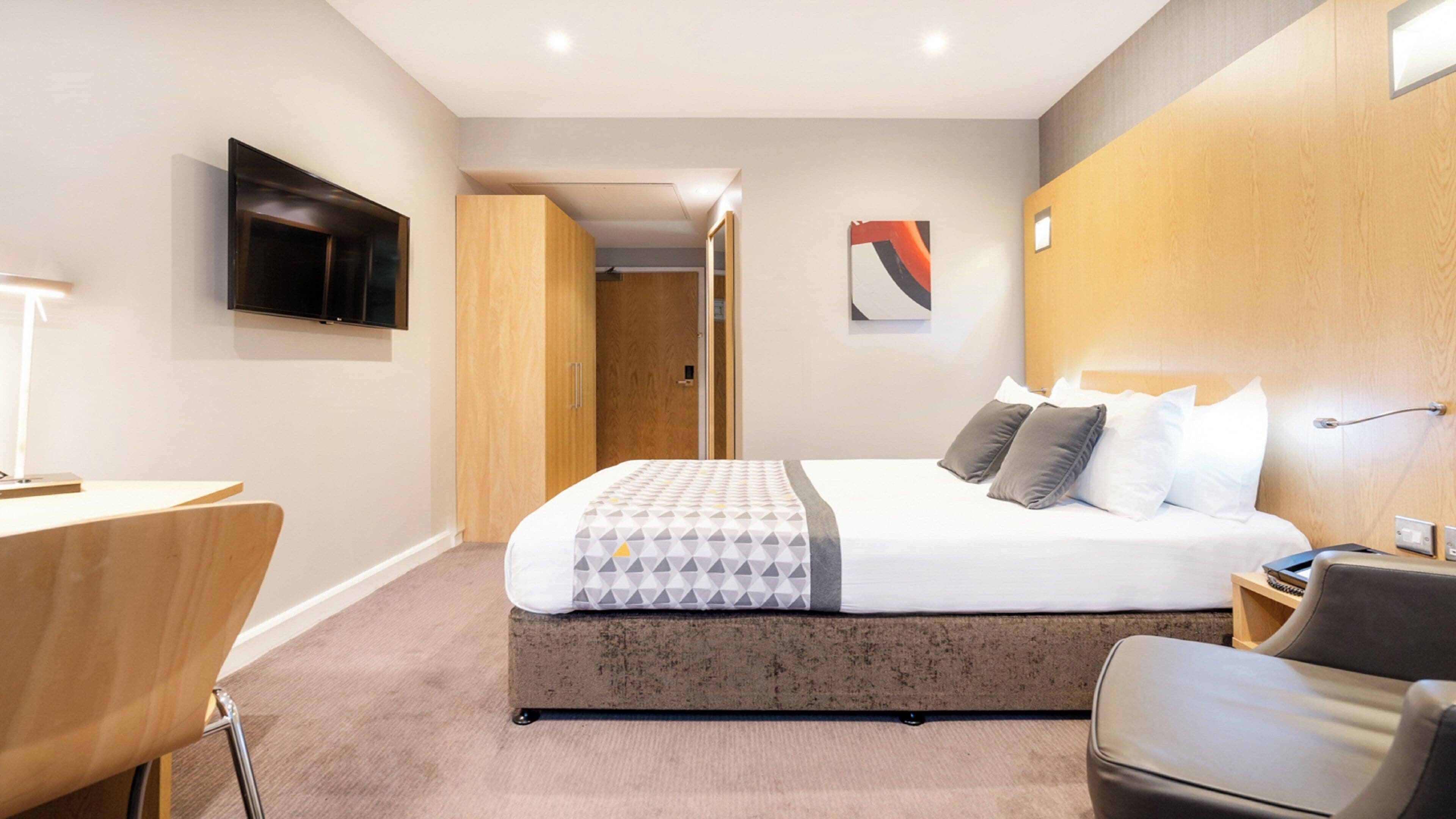 Quarto de hotel moderno com uma cama, uma escrivaninha, uma cadeira, uma televisão e uma porta que dá para outro espaço.