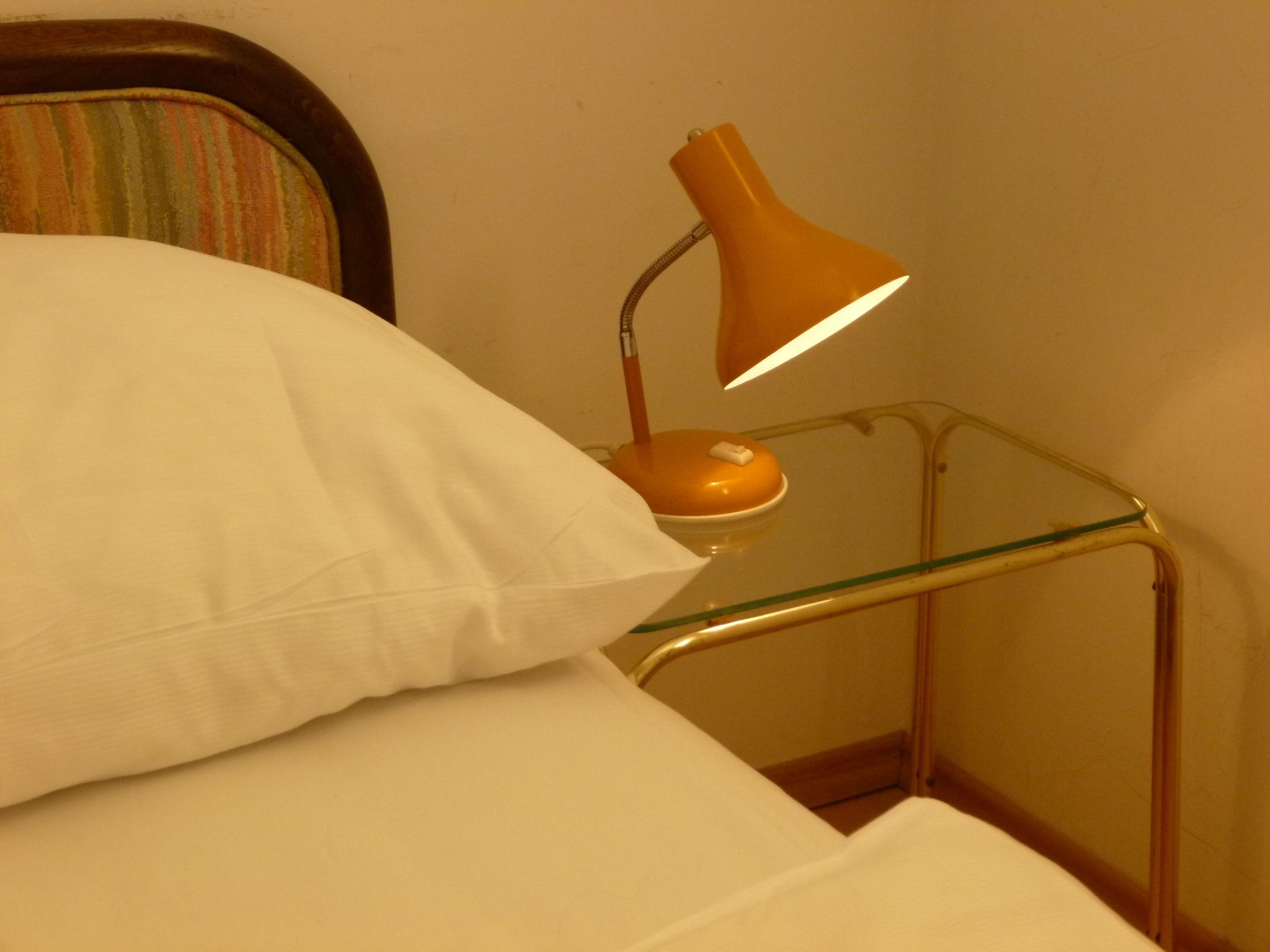 Uma cama com travesseiro branco e mesa de cabeceira de vidro com abajur amarelo.