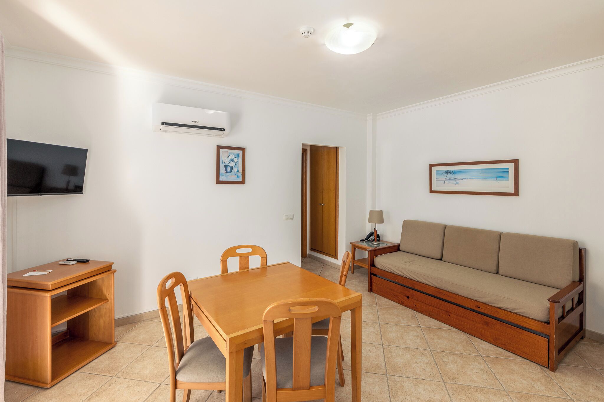 Um quarto com uma mesa de jantar de madeira, cadeiras, um sofá, uma televisão, um ar condicionado e um quadro pendurado na parede.