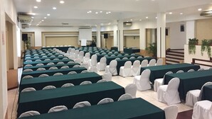 Uma sala de conferências com fileiras de mesas e cadeiras, equipada com projetor e alto-falantes.