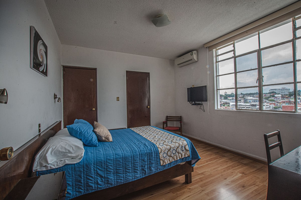 Quarto com cama, cadeira, escrivaninha e janela com vista para a cidade.