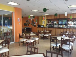Interior de restaurante com várias mesas e cadeiras, área de bar com televisão e quadros nas paredes.