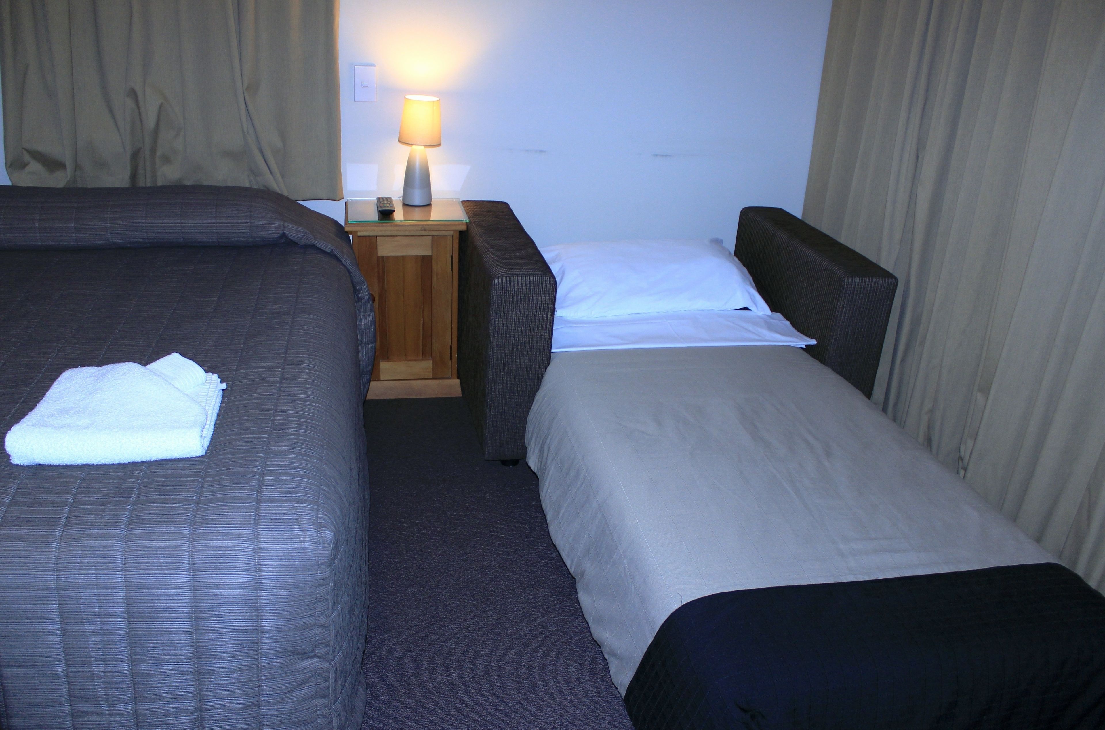 Quarto de hotel com duas camas de solteiro, uma mesa de cabeceira com abajur e um criado-mudo.