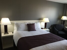 Quarto de hotel com uma cama grande, duas luminárias de cabeceira, uma cadeira e mesa de cabeceira com relógio.