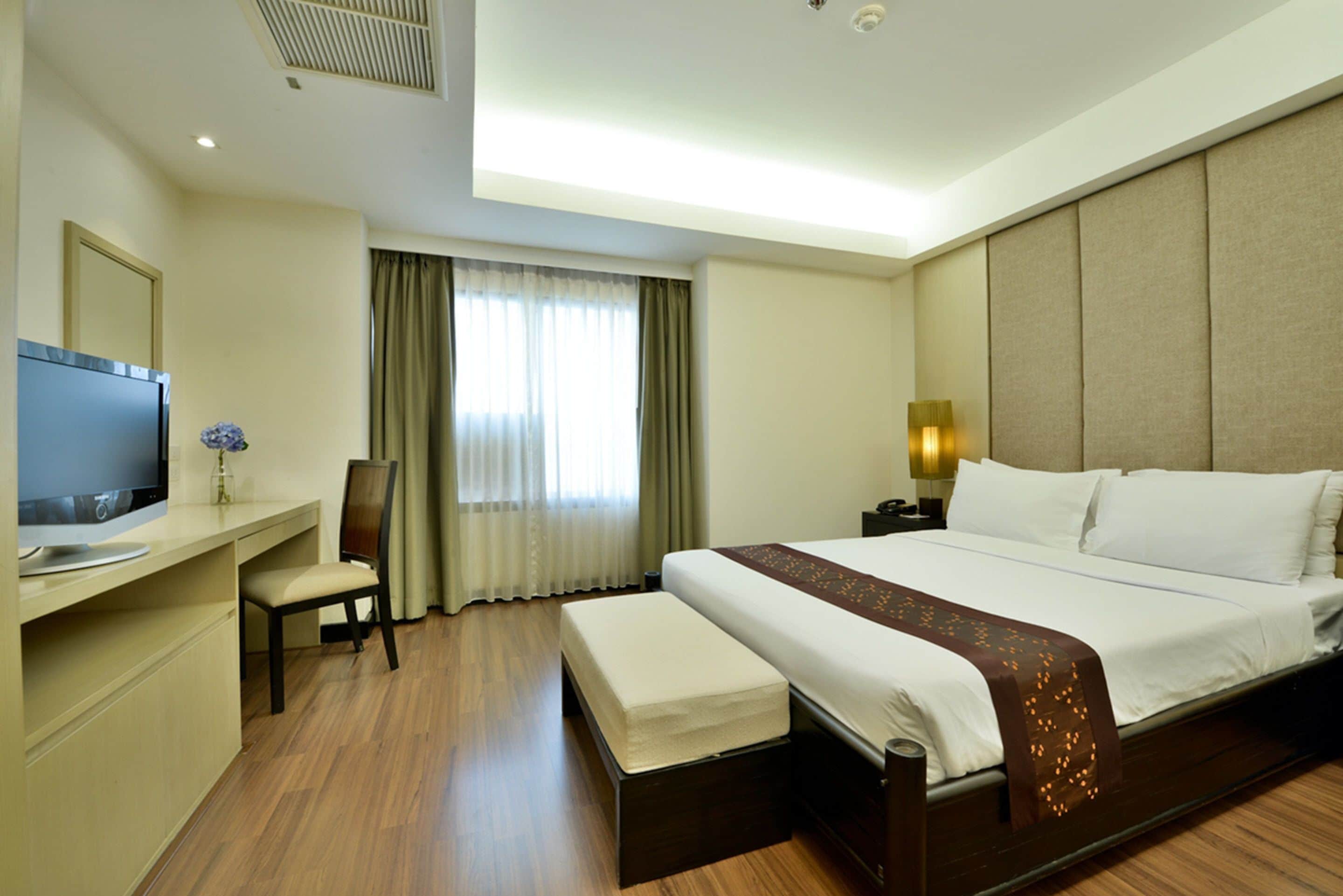 Quarto de hotel com cama, escrivaninha com televisão, cadeira, mesa de cabeceira com abajur e janela com cortinas.