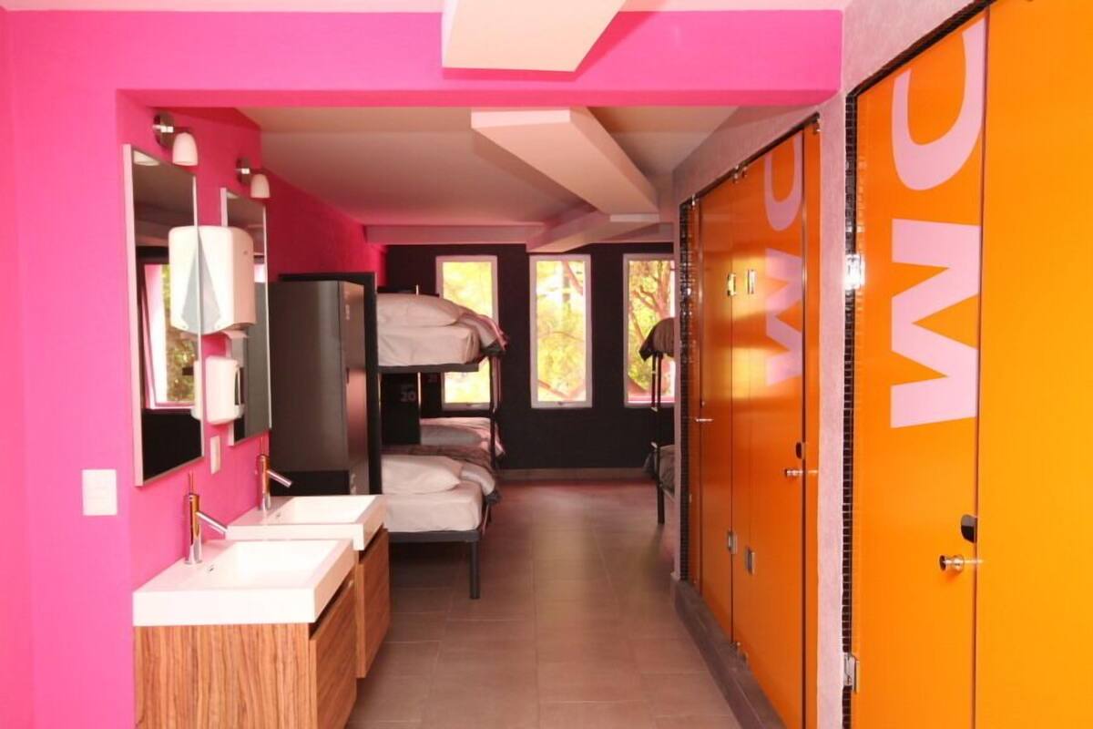 Quarto de hotel com paredes cor-de-rosa, toucador de madeira, duas pias, uma cama e um guarda-roupa com um grande logótipo 'M'.