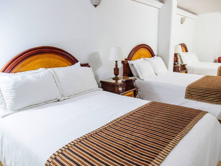 Quarto de hotel com duas camas, cada uma com roupa de cama branca e cabeceiras de madeira. Há mesas de cabeceira com abajures e um espelho.