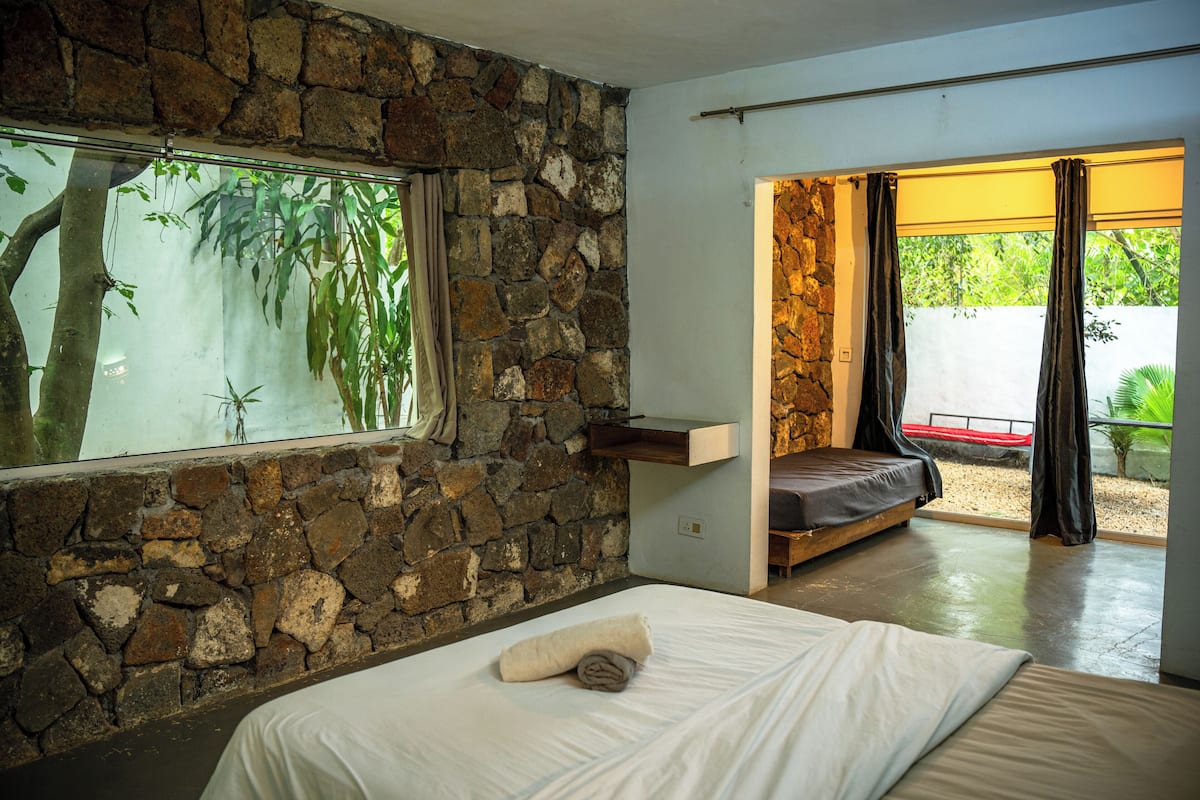 Quarto com parede de pedra, cama com roupa branca, janela com vista para vegetação e porta que dá para o exterior.