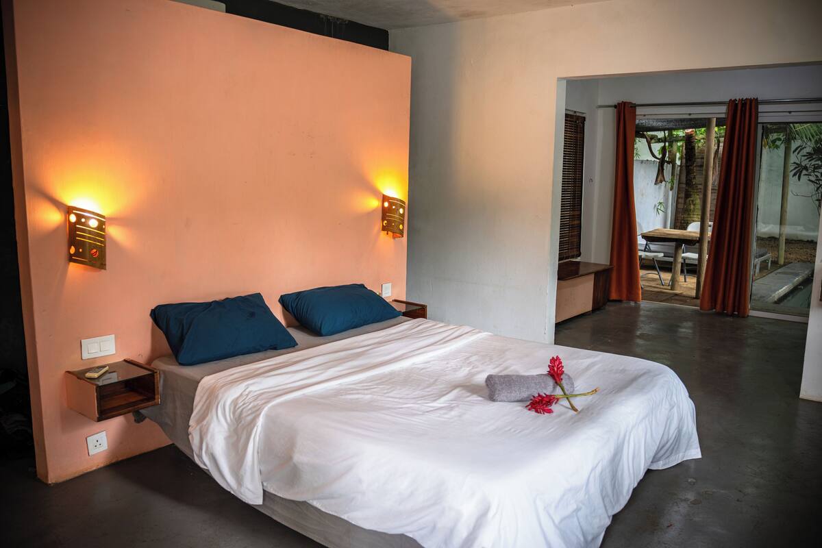Quarto de hotel moderno com uma cama grande, duas luminárias fixadas na parede, uma mesa de cabeceira e vista para o exterior por uma porta de vidro deslizante.