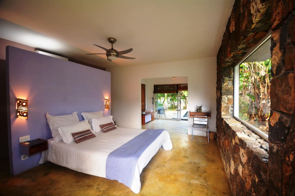 Quarto com cama, mesas de cabeceira, ventilador de teto e vista pela janela.