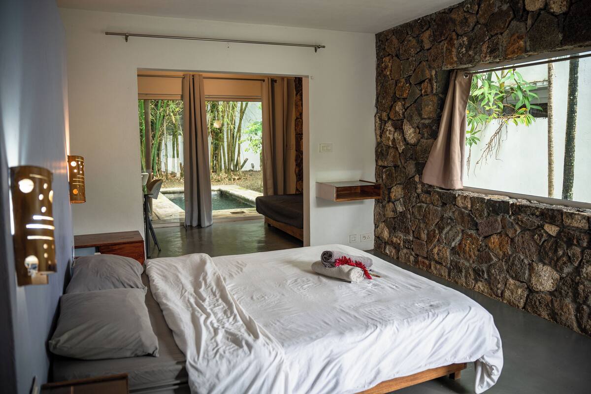 Quarto com parede de pedra, cama com roupa de cama branca, mesa de cabeceira de madeira e vista para um jardim pela janela.