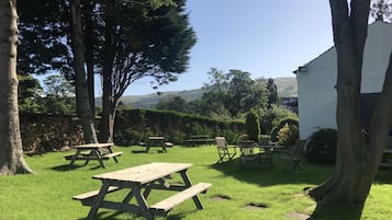 Uma área externa com gramado, mesas de piquenique e bancos, cercada por árvores e um edifício branco.