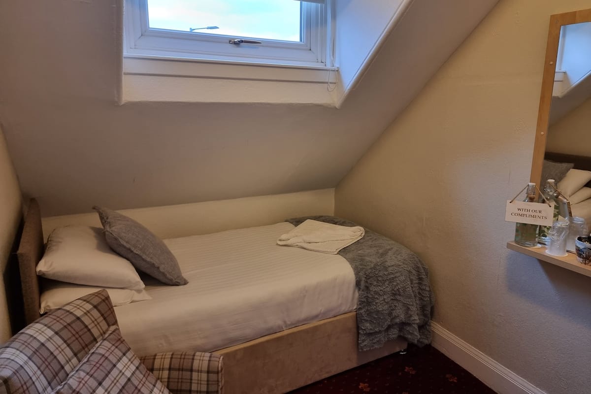 Quarto pequeno com cama de solteiro, teto inclinado e claraboia. Há uma mesa de cabeceira com um espelho e uma prateleira com artigos de higiene pessoal.