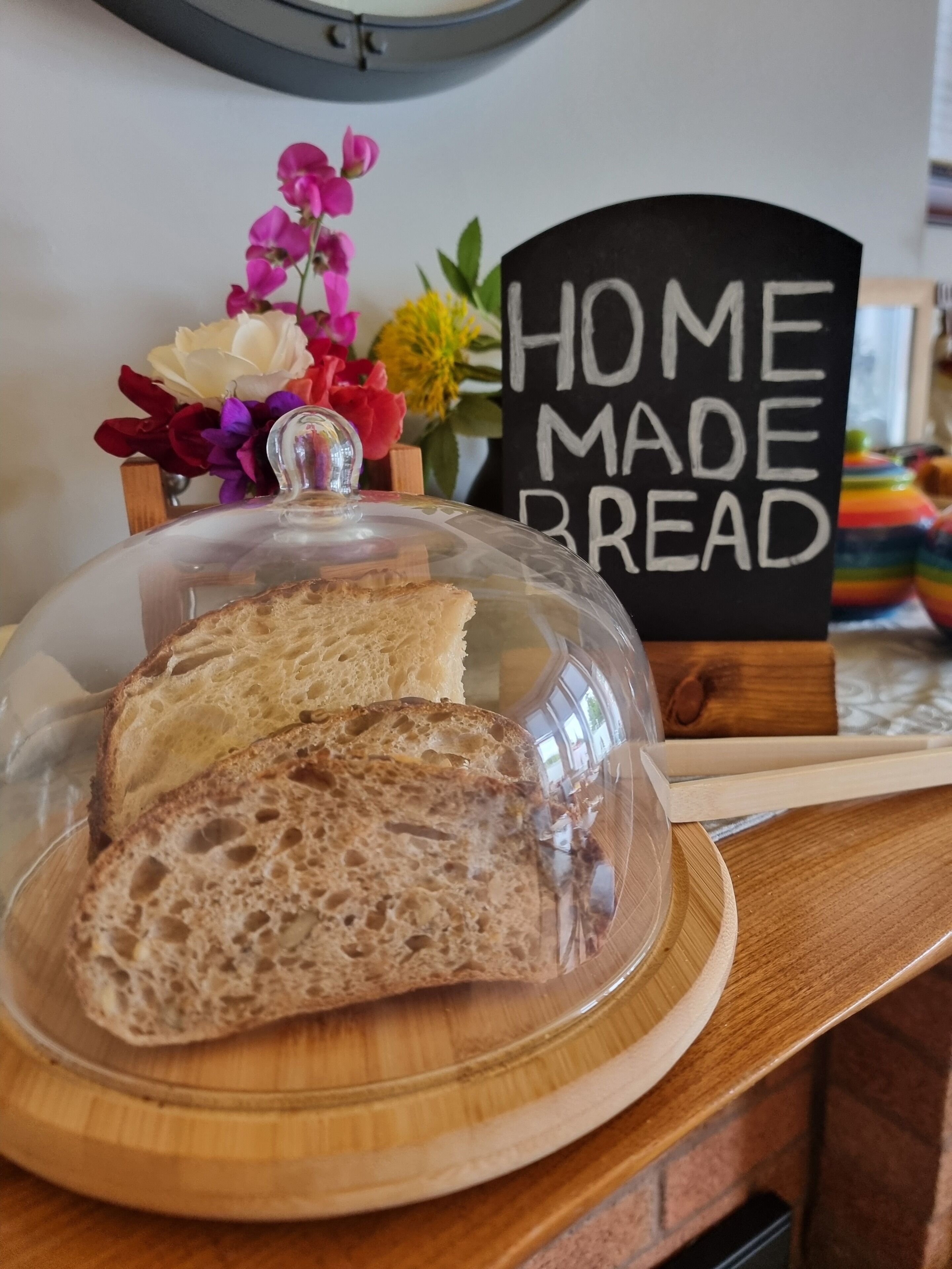 Um balcão de madeira com uma cúpula de vidro exibindo fatias de pão caseiro, um letreiro com a inscrição "HOME MADE BREAD" e um vaso de flores ao fundo.