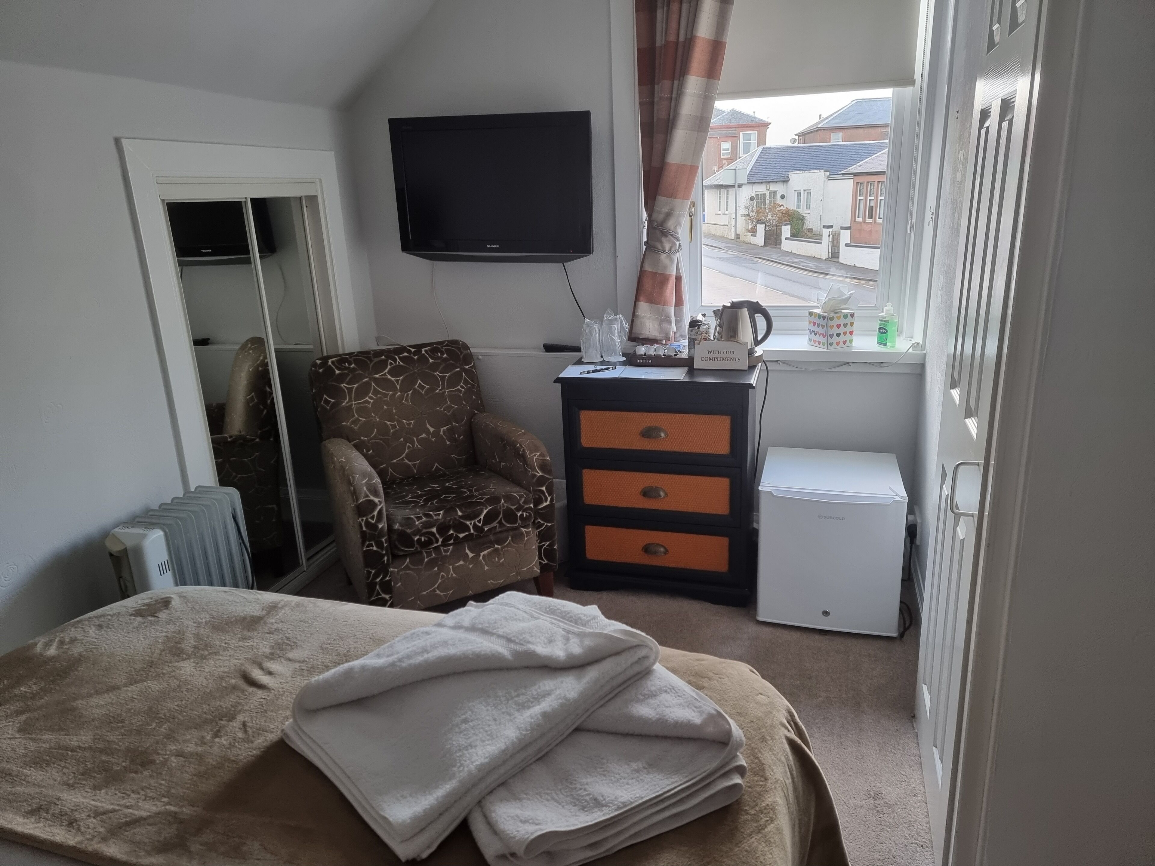 Um quarto com cama, cadeira, cômoda, televisão e janela com vista para edifícios.