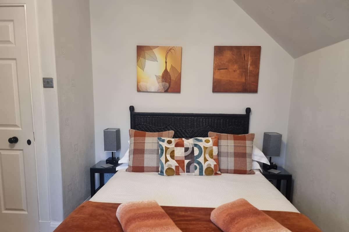 Cama bem arrumada com duas toalhas, cabeceira preta e duas luminárias fixadas nas paredes, uma de cada lado.