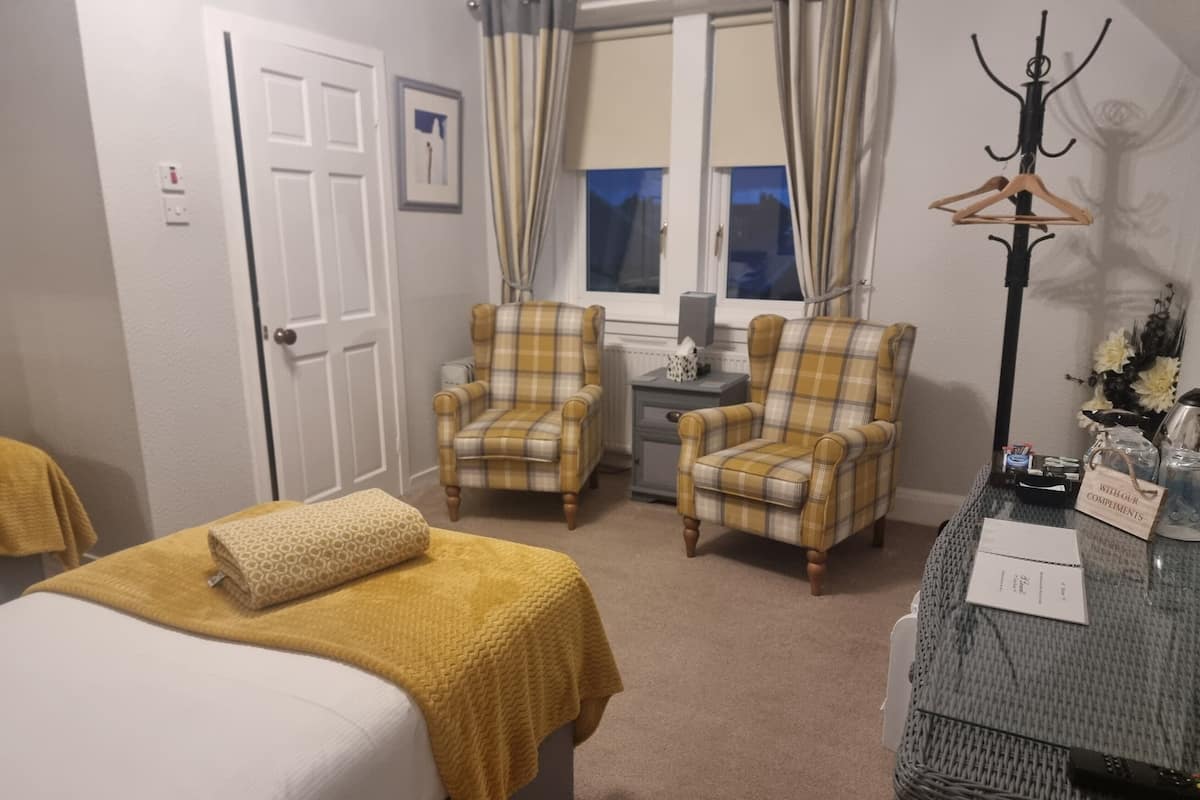 Quarto de hotel com duas poltronas xadrez, uma cama com manta amarela, uma mesinha com caixa de lenços e um cabideiro com ganchos.