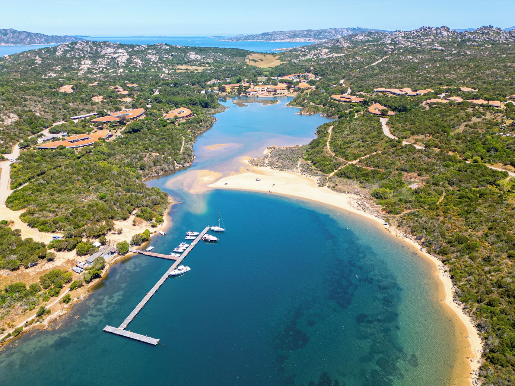 Vista aérea de uma área costeira com um porto de iates, praia de areia e vegetação ao redor.