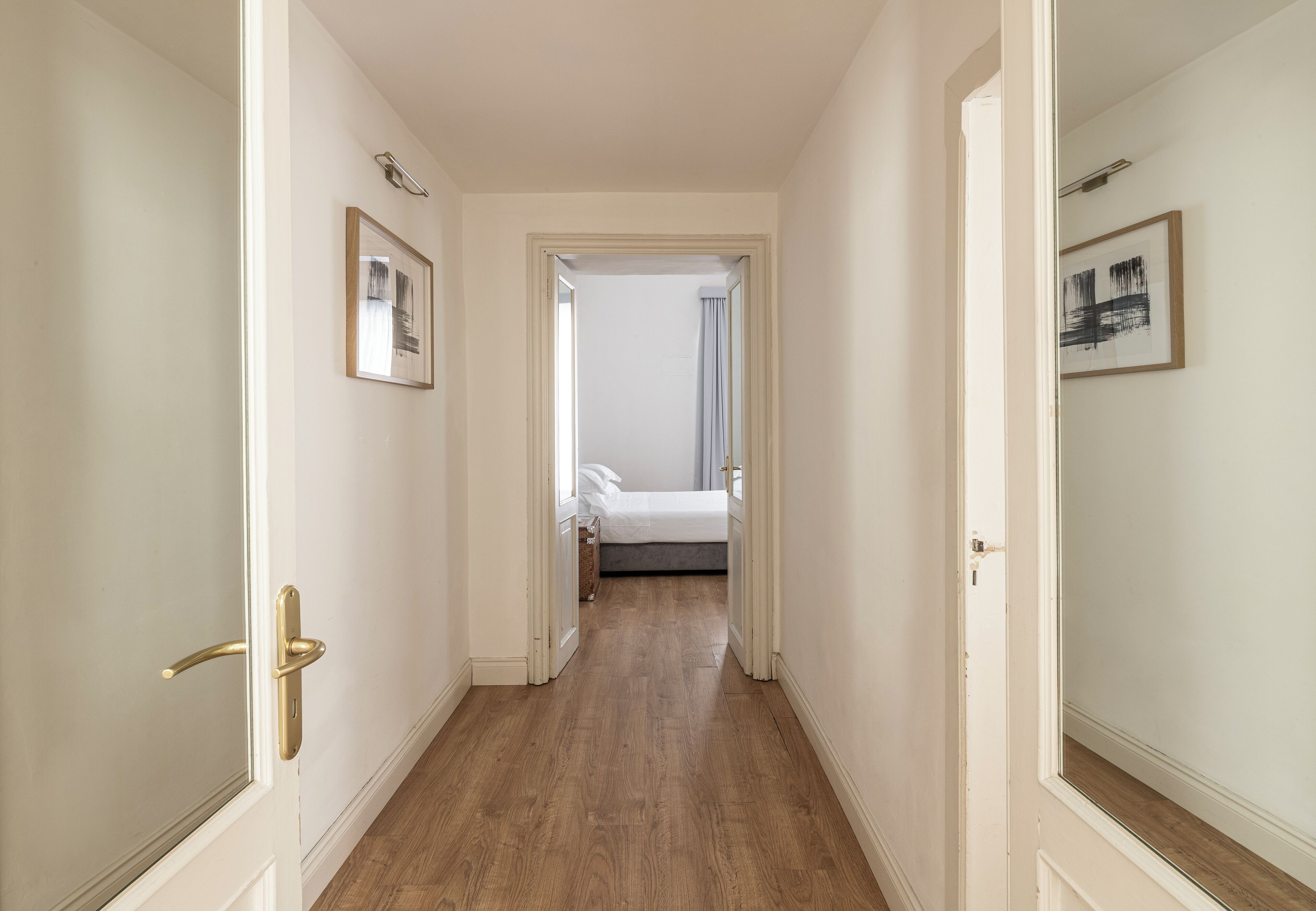 Um corredor estreito com piso de madeira e paredes brancas. Há quadros nas paredes, uma porta com puxador de latão e uma cama com cabeceira branca ao fundo.