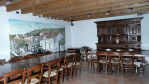 Uma sala de jantar com uma mesa de madeira comprida rodeada por cadeiras, um quadro grande na parede e um armário de madeira com esculturas detalhadas.