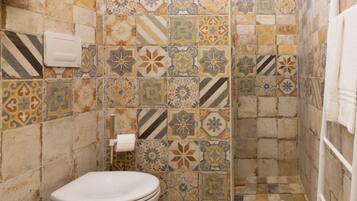 Casa de banho com sanita, área de duche e parede revestida a mosaicos com diferentes padrões.
