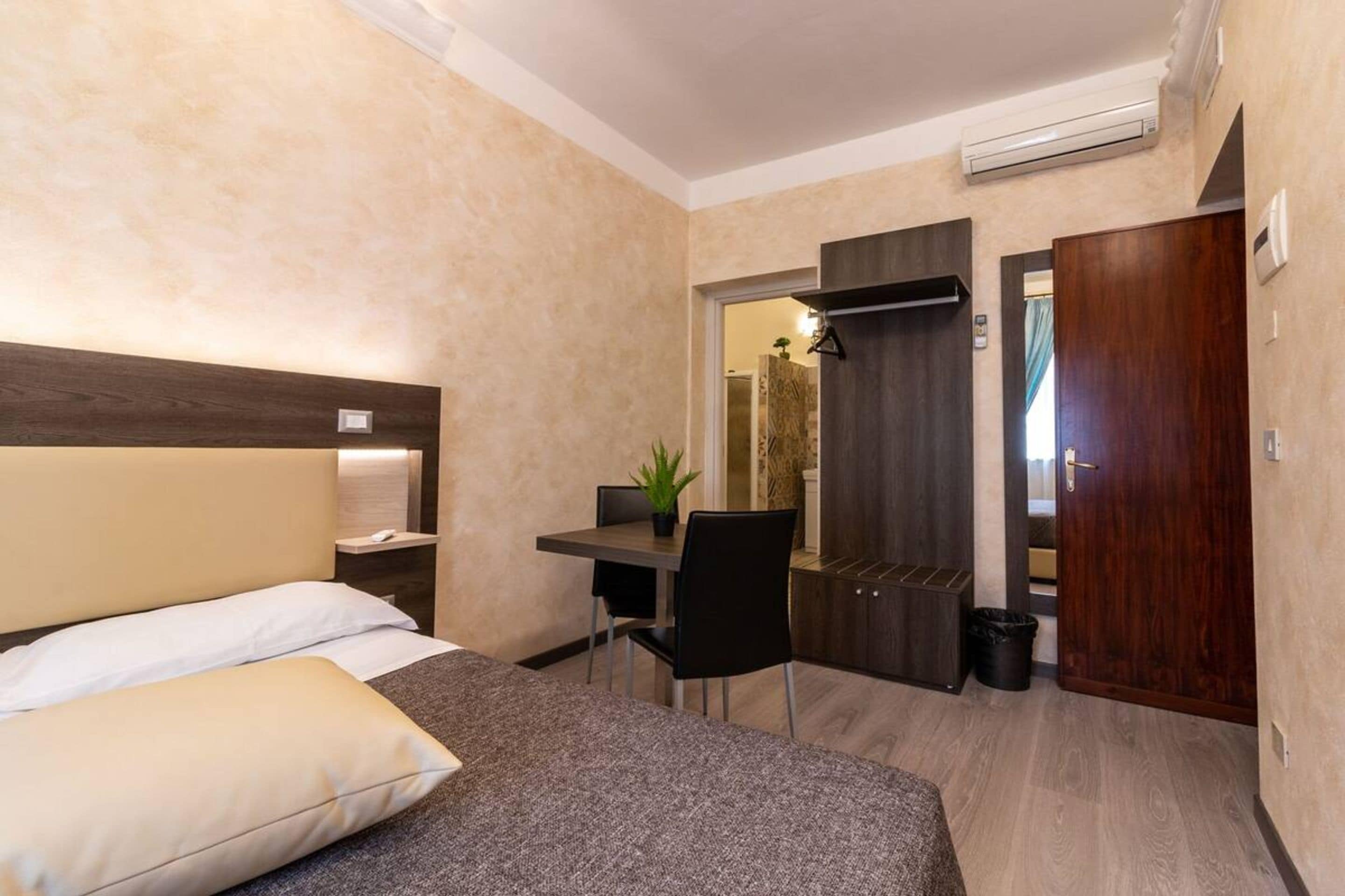 Quarto de hotel com uma cama, escrivaninha, cadeira, espelho e guarda-roupa.
