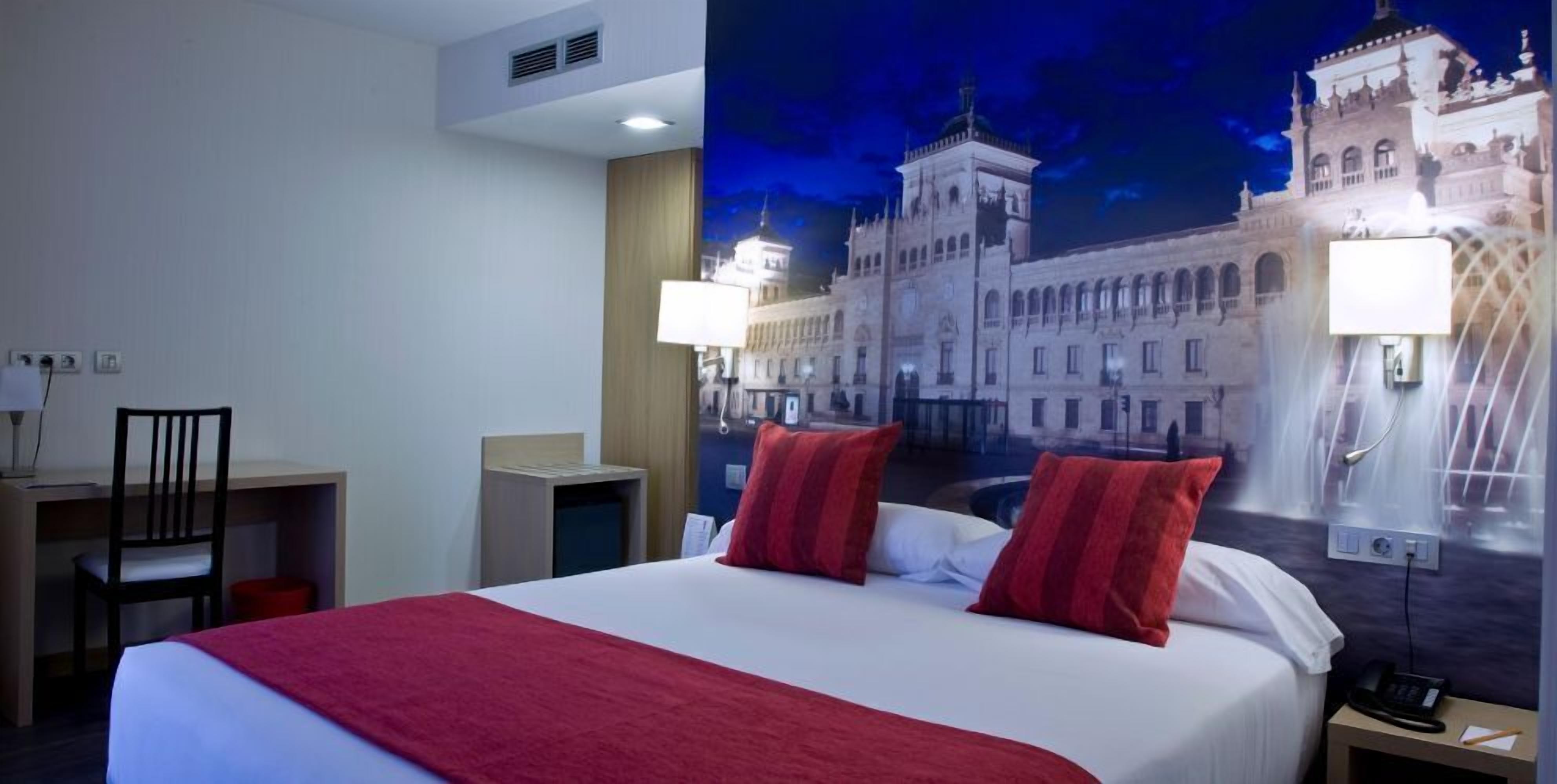 Quarto de hotel com cama, escrivaninha, cadeira e mesa de cabeceira com abajur.