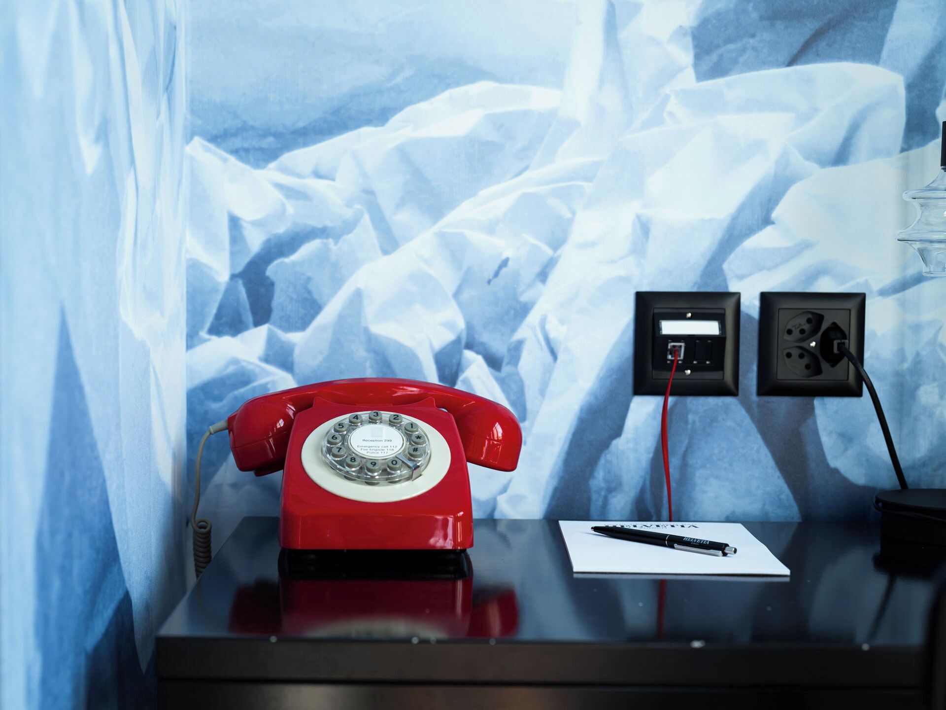 Um telefone rotativo vermelho sobre uma mesa, com um papel branco e uma caneta, em frente a uma parede com textura de papel amassado.