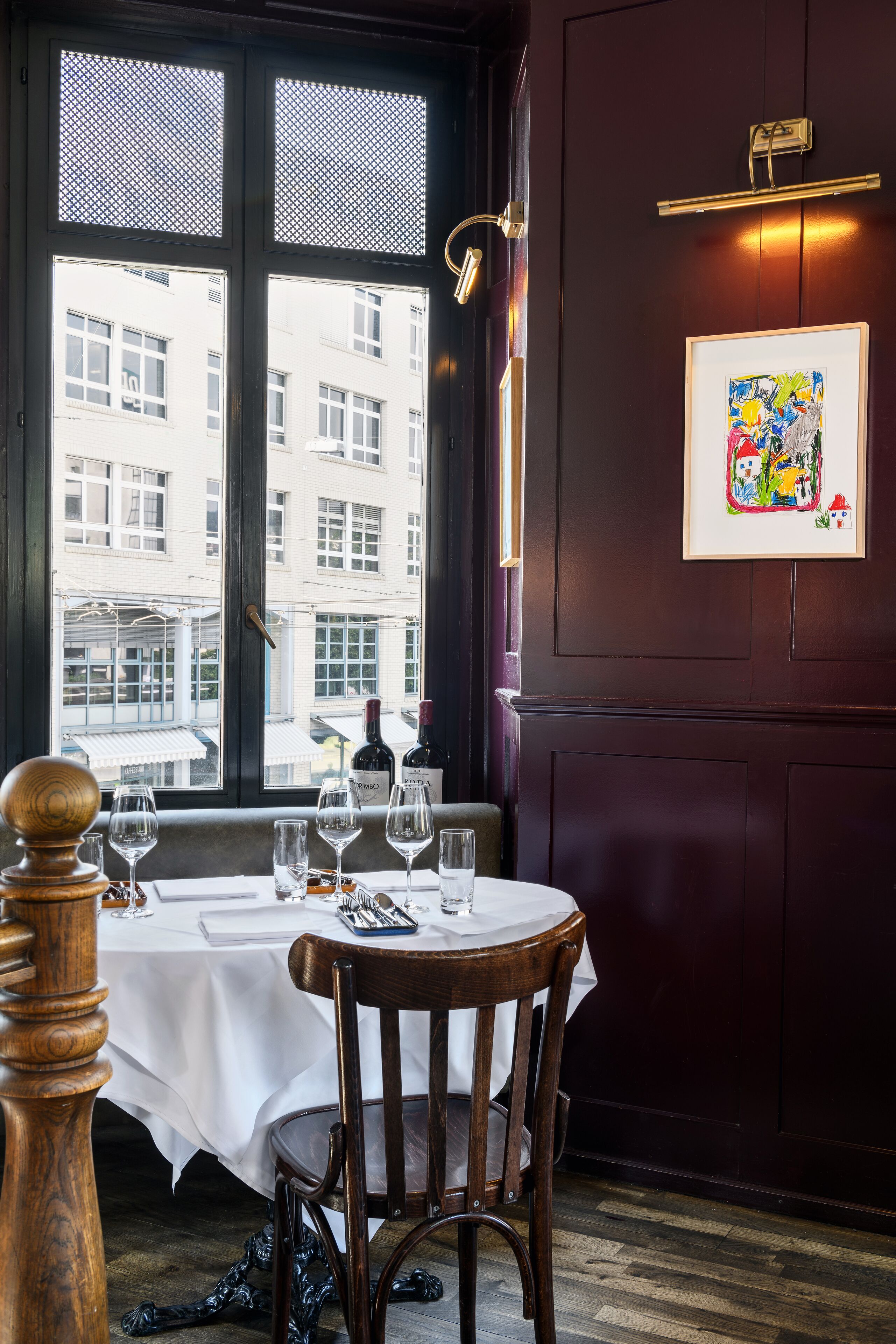 Uma área de refeições com uma mesa posta para dois, uma janela com vista para edifícios e uma obra de arte emoldurada na parede.