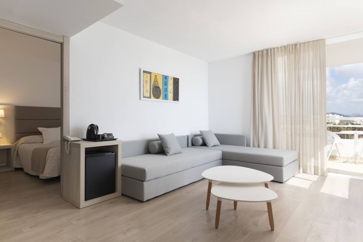 Quarto de hotel moderno com sofá cinzento, uma mesa pequena, secretária com máquina de café e vista para a cidade através de janelas amplas.