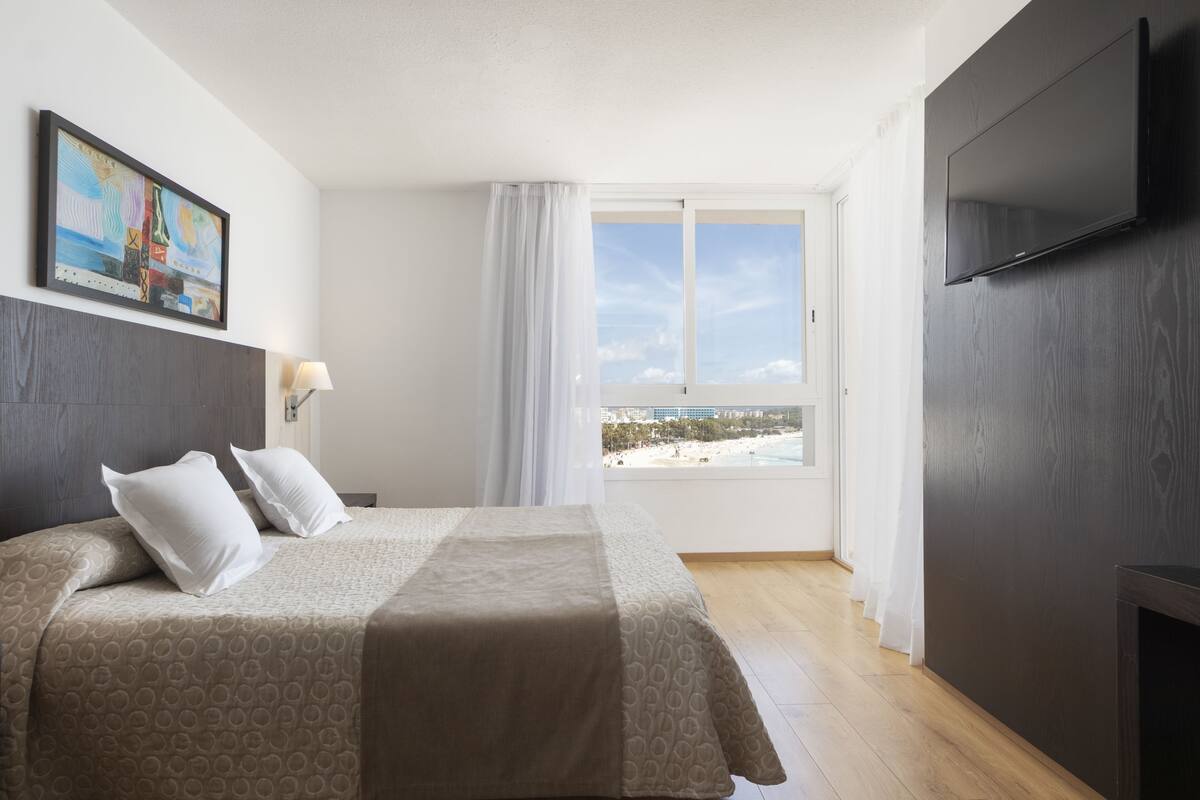 Quarto de hotel com cama, televisão, pintura na parede e janela vista para a praia.