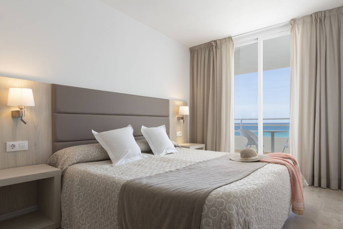 Quarto de hotel com uma cama grande, duas luminárias de cabeceira, uma mesinha de cabeceira, um chapéu e vista para o mar através de uma varanda.