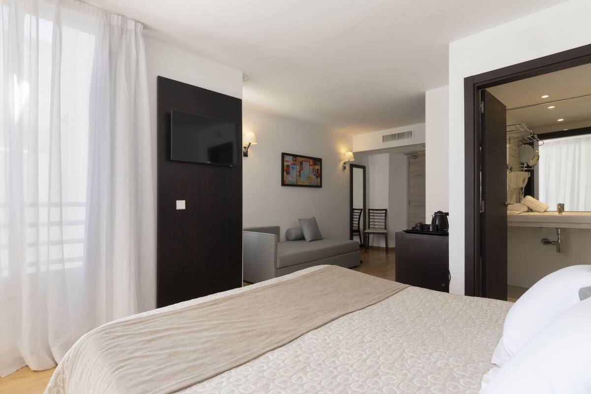 Quarto de hotel com cama, sofá, televisão, um quadro e banheiro visível por uma porta aberta.