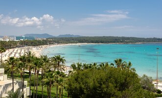 Uma praia com águas turquesa cristalinas, palmeiras e um hotel ao fundo.
