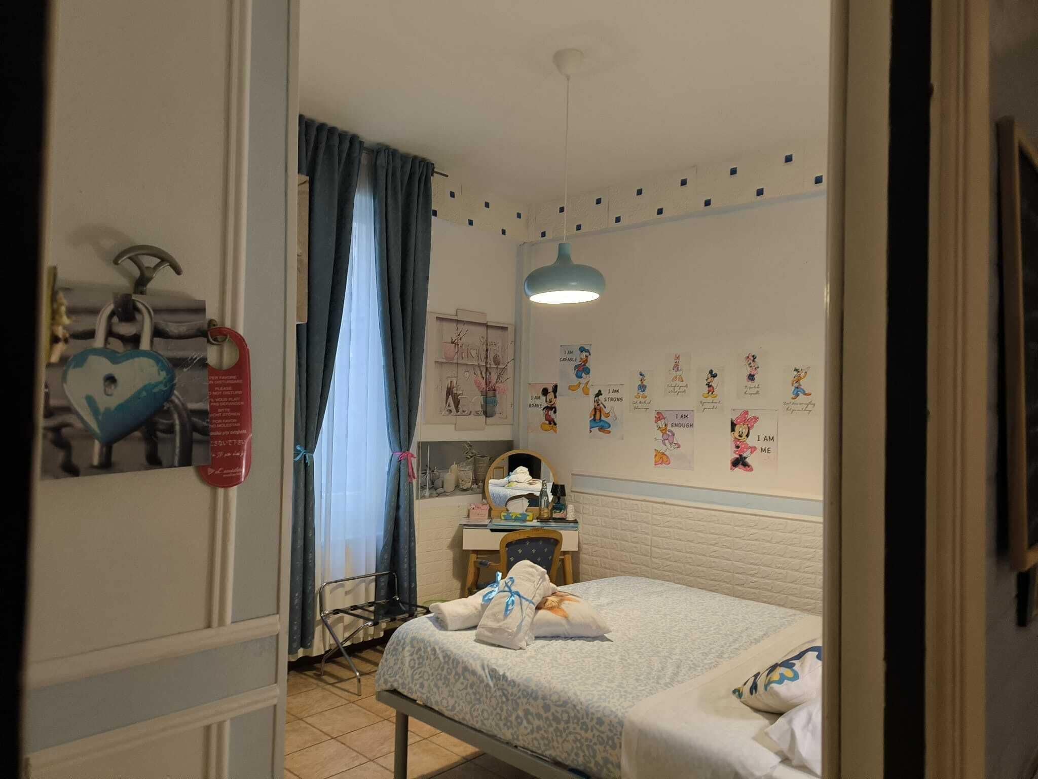 Quarto com cama, escrivaninha e cadeira. Cortinas, relógio de parede e cartazes motivacionais.