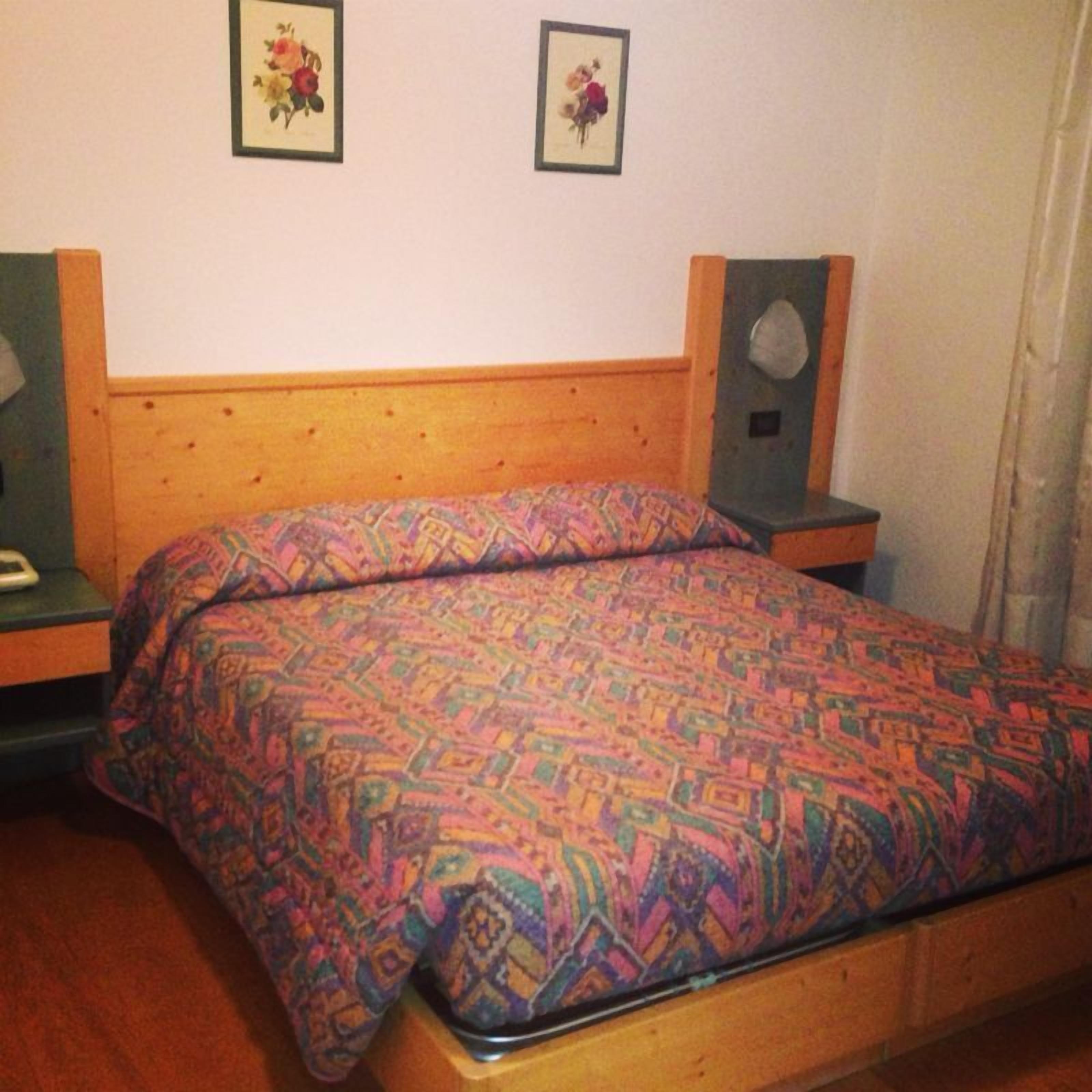 Uma cama de solteiro com colcha estampada, duas mesas de cabeceira com abajures, e quadros florais na parede.