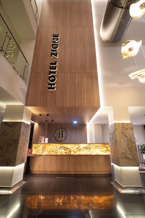 Uma área de recepção de hotel com um balcão de madeira, uma parede de destaque em mármore e um candelabro moderno.